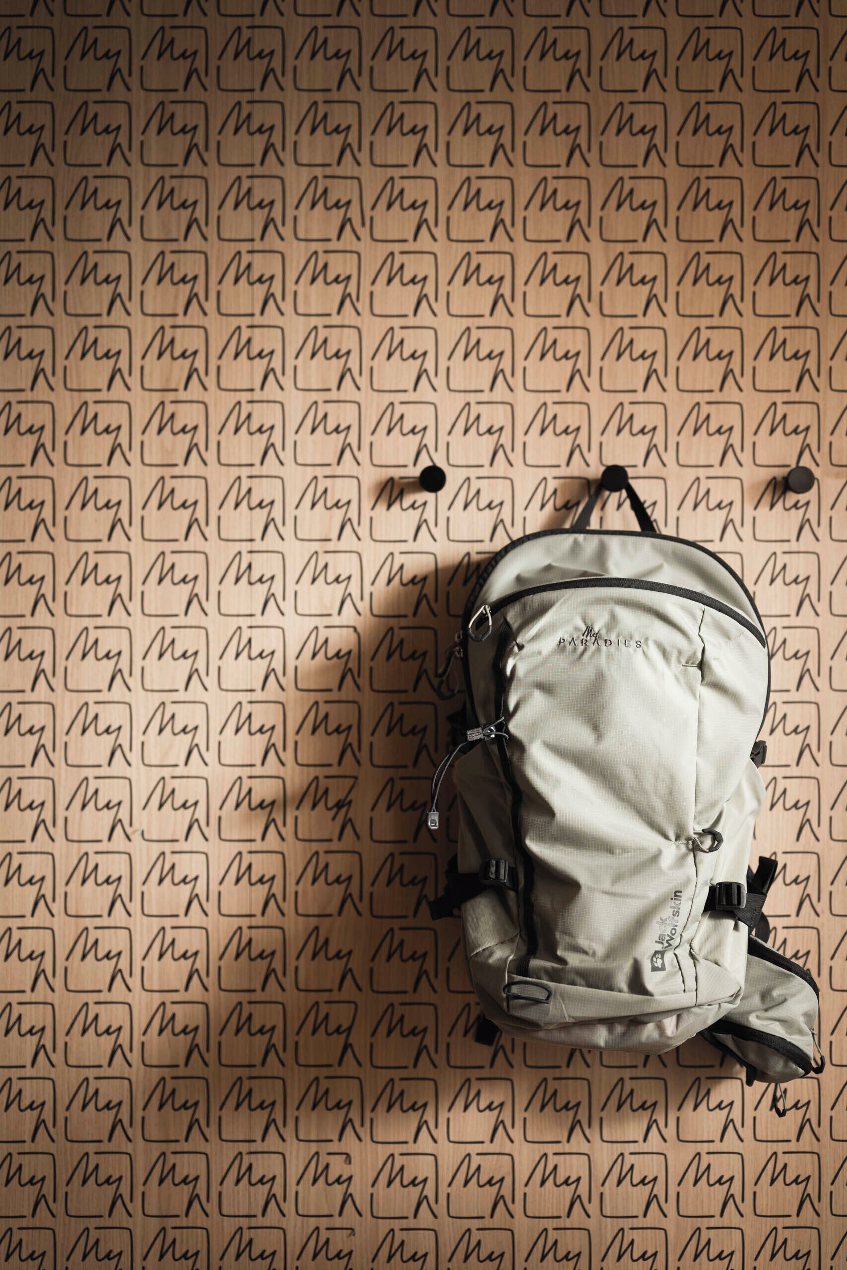 Mochila com a palavra 'PARADISE' escrita nela, pendurada em uma parede com um padrão repetitivo da palavra 'My'.