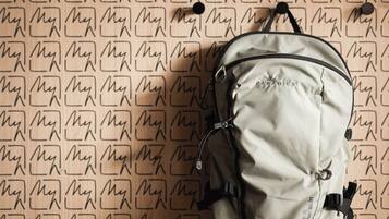 Mochila com a palavra 'PARADISE' escrita nela, pendurada em uma parede com um padrão repetitivo da palavra 'My'.
