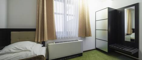 Quarto de hotel com cama, radiador, janela com cortinas, guarda-roupa e espelho.
