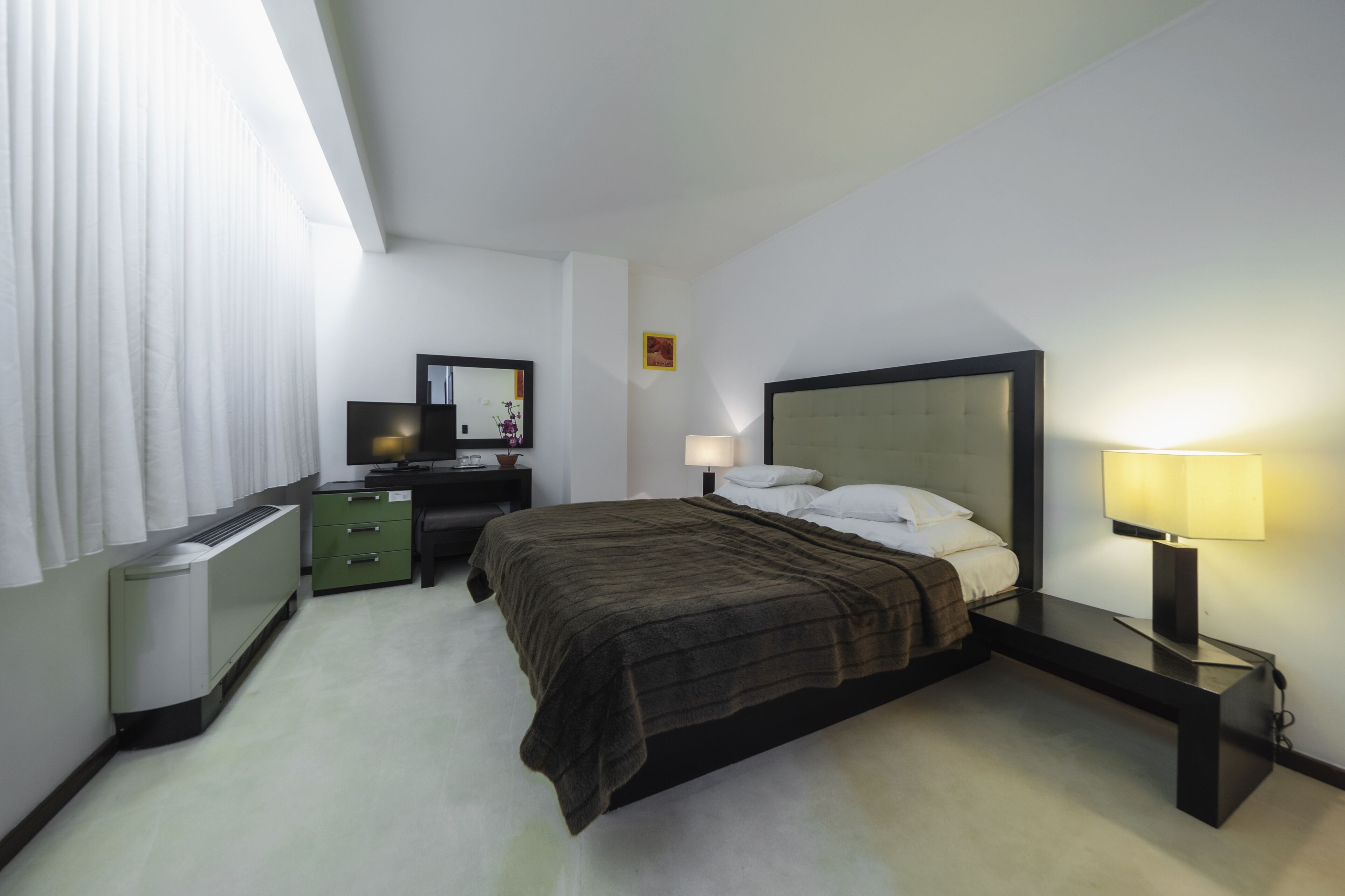Quarto de hotel com cama, mesa de cabeceira com luminária, cômoda, televis ão e radiador.