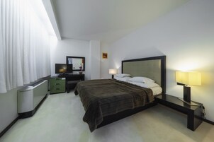 Quarto de hotel com cama, mesa de cabeceira com luminária, cômoda, televisão e radiador.