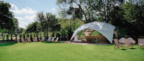 Uma área de estar externa com estrutura de toldo branco, cercada por árvores e um gramado.