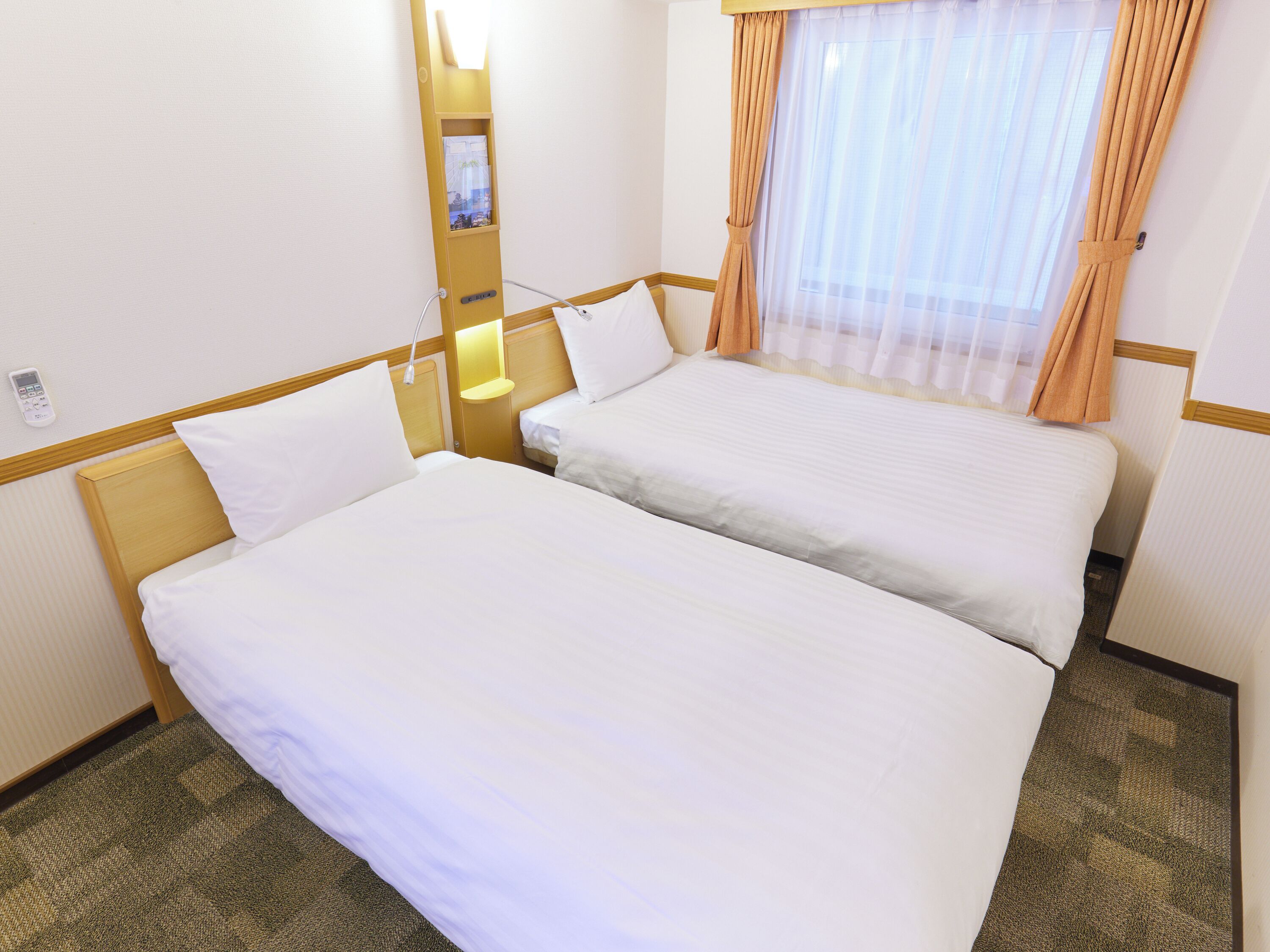 Quarto de hotel com duas camas, uma mesa de cabeceira com abajur, uma janela com cortinas e um telefone fixado na parede.