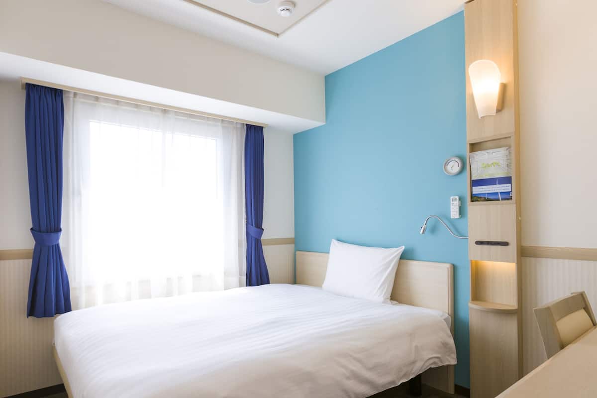 Quarto de hotel com uma cama grande, uma escrivaninha, uma cadeira, um relógio fixado na parede e um painel iluminado.