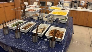 Disposição de buffet com diversos alimentos em bandejas, um espaço para café e uma cozinha ao fundo.