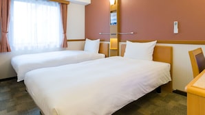 Quarto de hotel com duas camas, uma janela grande com cortinas, uma luminária na parede e uma pequena escrivaninha.