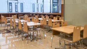 Uma sala de aula com mesas e cadeiras de madeira, janelas amplas e um quadro branco.
