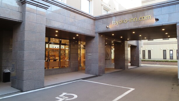 Entrada de um prédio com um letreiro 'toyoko-in.com', fachada de vidro e um espaço para estacionamento de bicicletas.