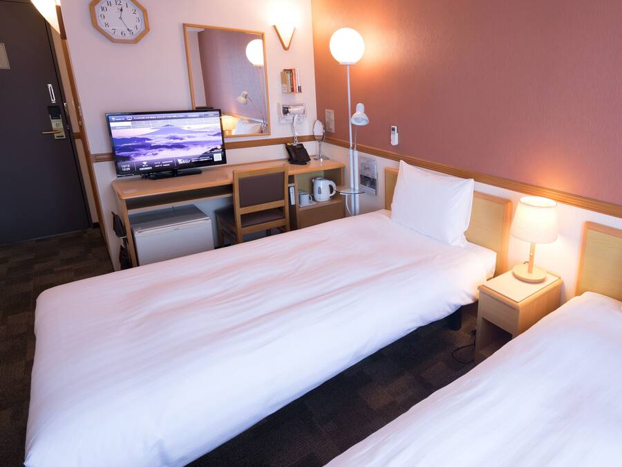 Quarto de hotel com uma cama, uma escrivaninha com computador, uma cadeira, um abajur e um relógio na parede.