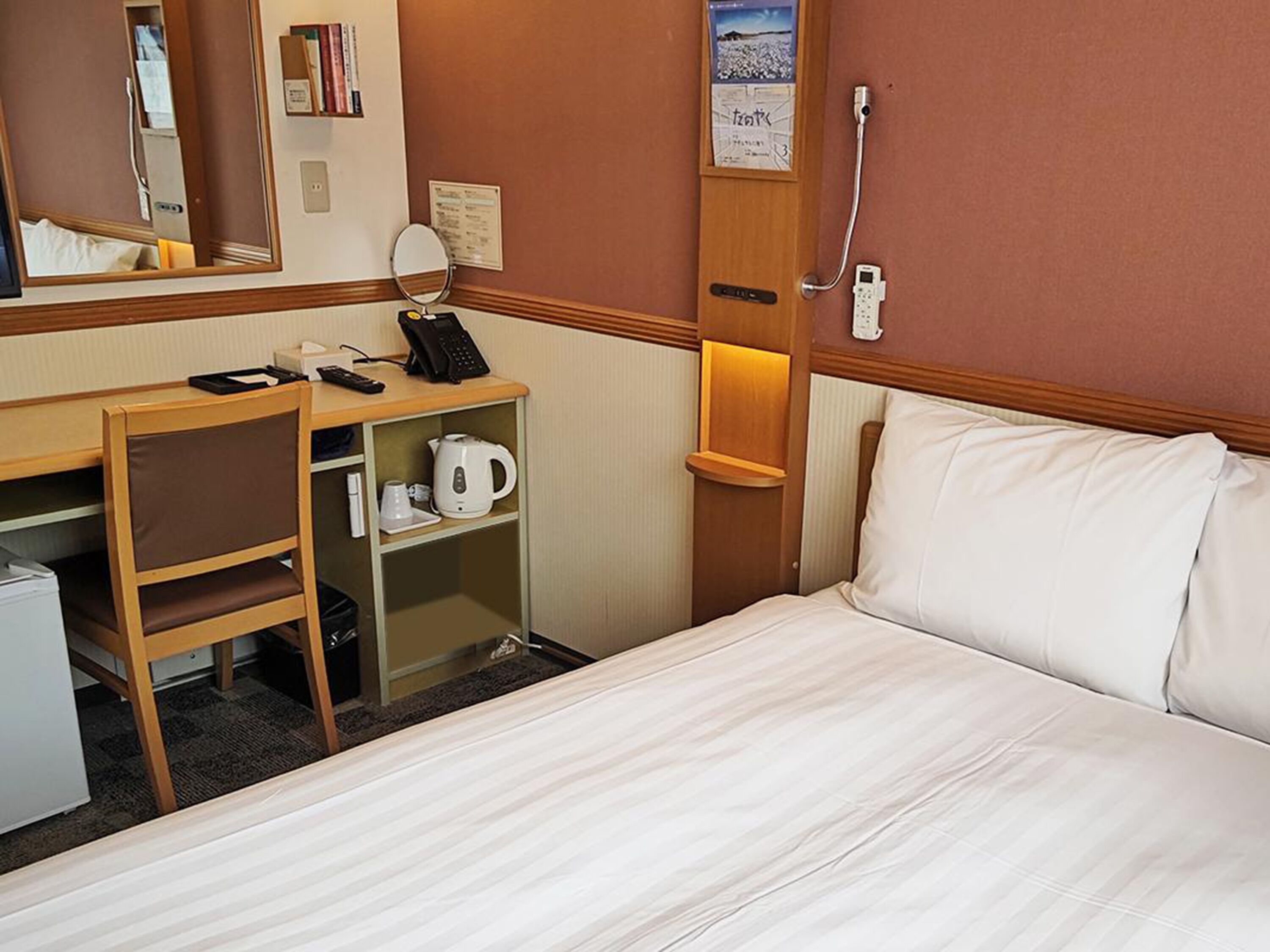 Quarto de hotel com cama, escrivaninha com telefone e chaleira, espelho e chuveiro.