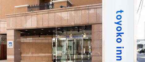 Exterior do Toyoko Inn, com entrada visível, placa de identificação e calçada de azulejos.