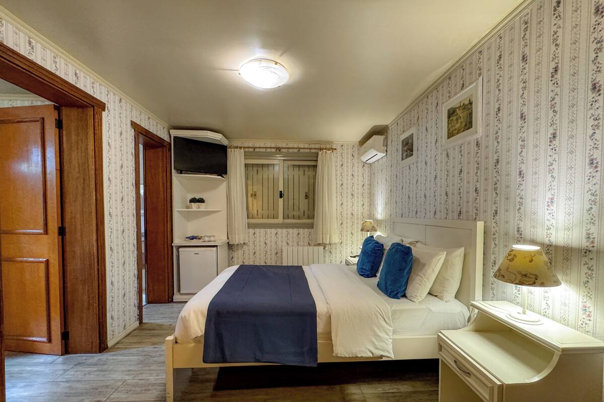 Quarto de hotel com cama, mesa de cabeceira e pequena escrivaninha com abajur.