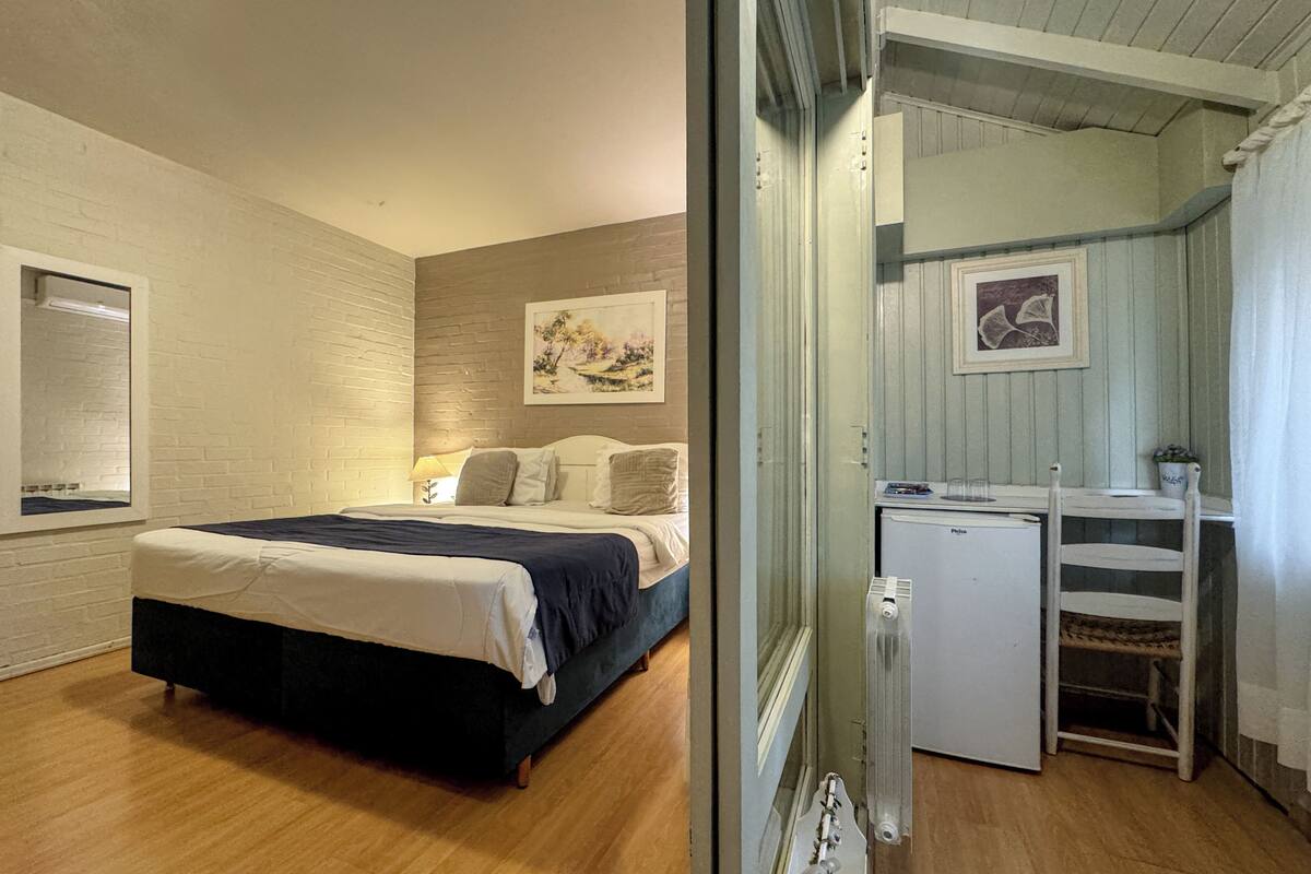 Quarto com cama, mesa de cabeceira, abajur e um pequeno frigorífico.