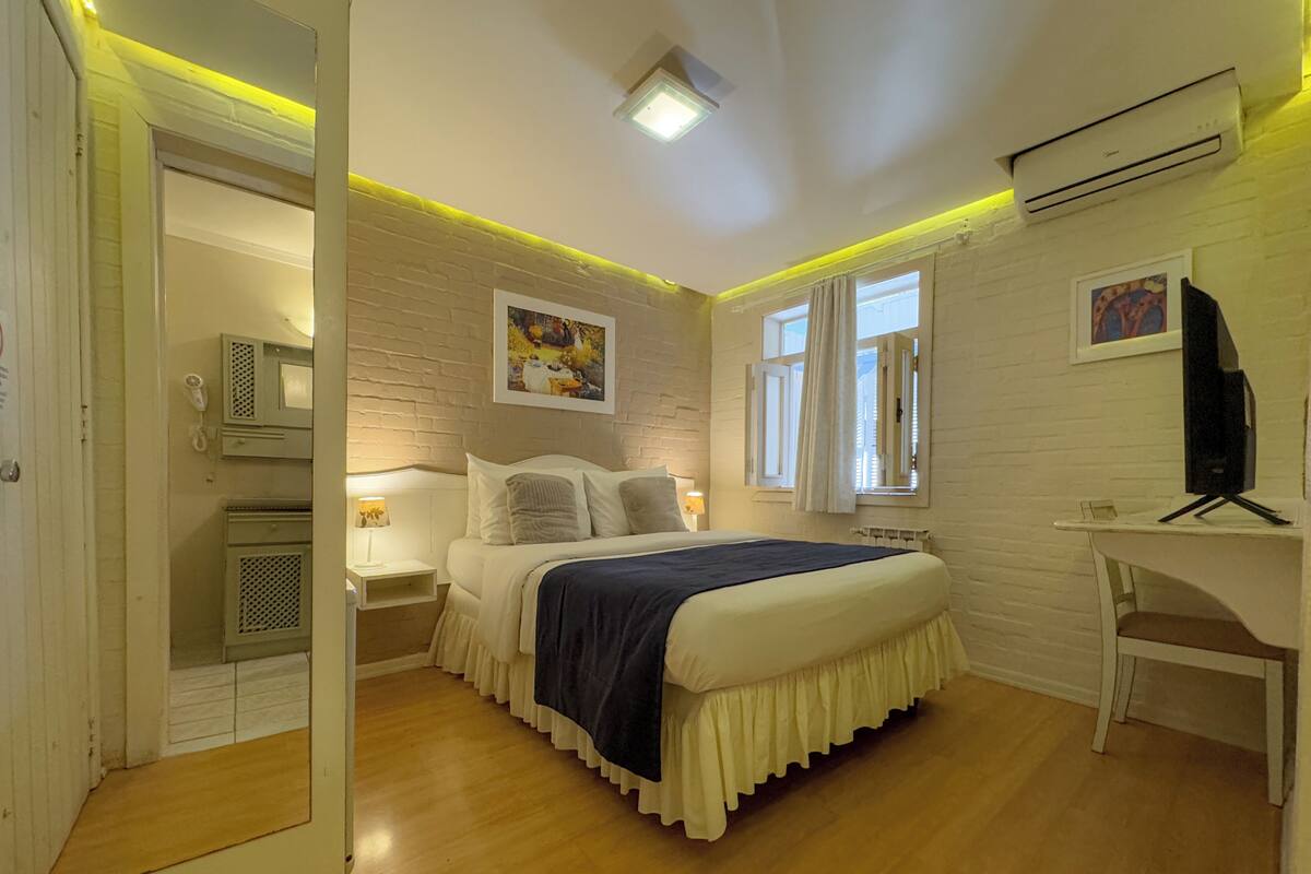 Quarto com cama, mesas de cabeceira, escrivaninha, ar condicionado e janela.