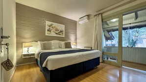 Quarto com cama, duas mesas de cabeceira com abajures, um quadro na parede e uma porta de vidro deslizante para uma varanda.