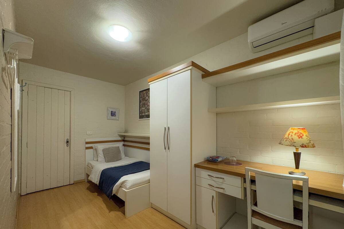 Quarto compacto com uma cama de solteiro, uma escrivaninha e uma cadeira. Há um guarda-roupa, uma pequena estante e um ar condicionado sobre a escrivaninha.