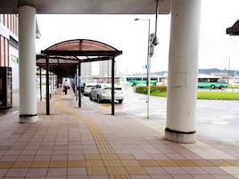 Uma passarela coberta para pedestres com piso tátil, um ponto de ônibus e uma fileira de carros estacionados.