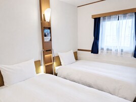 Quarto de hotel com duas camas, mesa de cabeceira, prateleira embutida com material de leitura e janela com cortinas.
