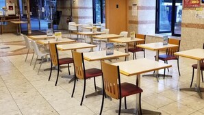 Uma cafeteria com várias mesas e cadeiras, piso de azulejos e vista para o exterior por grandes janelas.