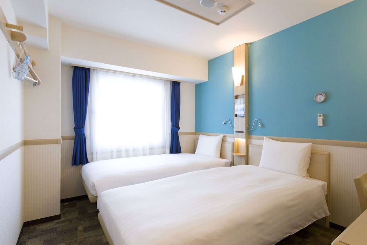 Quarto de hotel com duas camas, uma janela com cortinas, uma mesinha de cabeceira, um abajur e um relógio de parede.