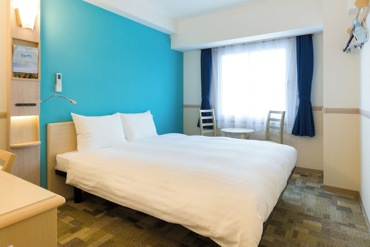 Quarto de hotel com uma cama grande, uma mesinha de cabeceira, uma escrivaninha pequena, uma cadeira, uma janela com cortinas e uma luminária na parede.