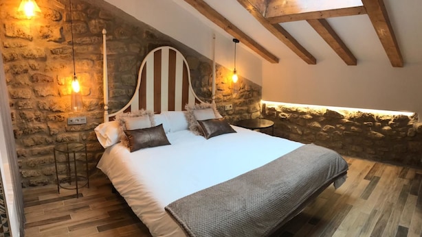 Quarto com parede de pedra, vigas de madeira, cama com almofadas e mesa de cabeceira.