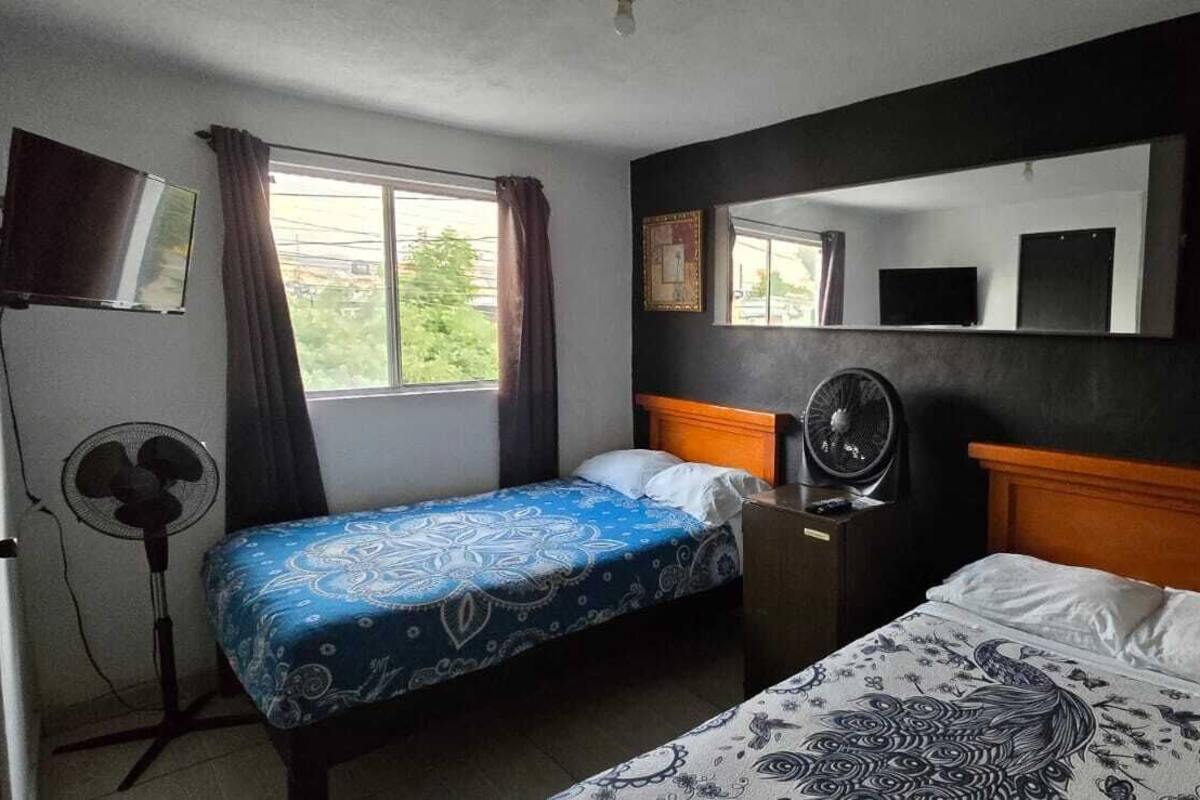Quarto com duas camas, televisão, ventoinha e janela com cortinas.