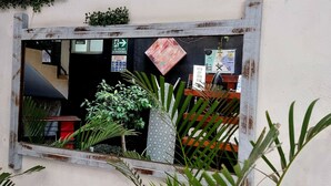 Janela com vista para um quarto que inclui cama, mesa de cabeceira e espelho. Plantas e uma porta são visíveis pela janela.
