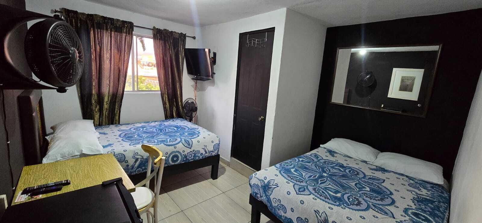 Quarto com duas camas, uma escrivaninha, uma cadeira, televisão e janela com cortinas.