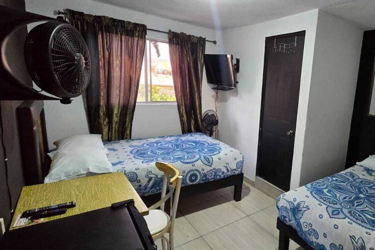 Quarto com duas camas, uma escrivaninha, uma cadeira, televisão e janela com cortinas.
