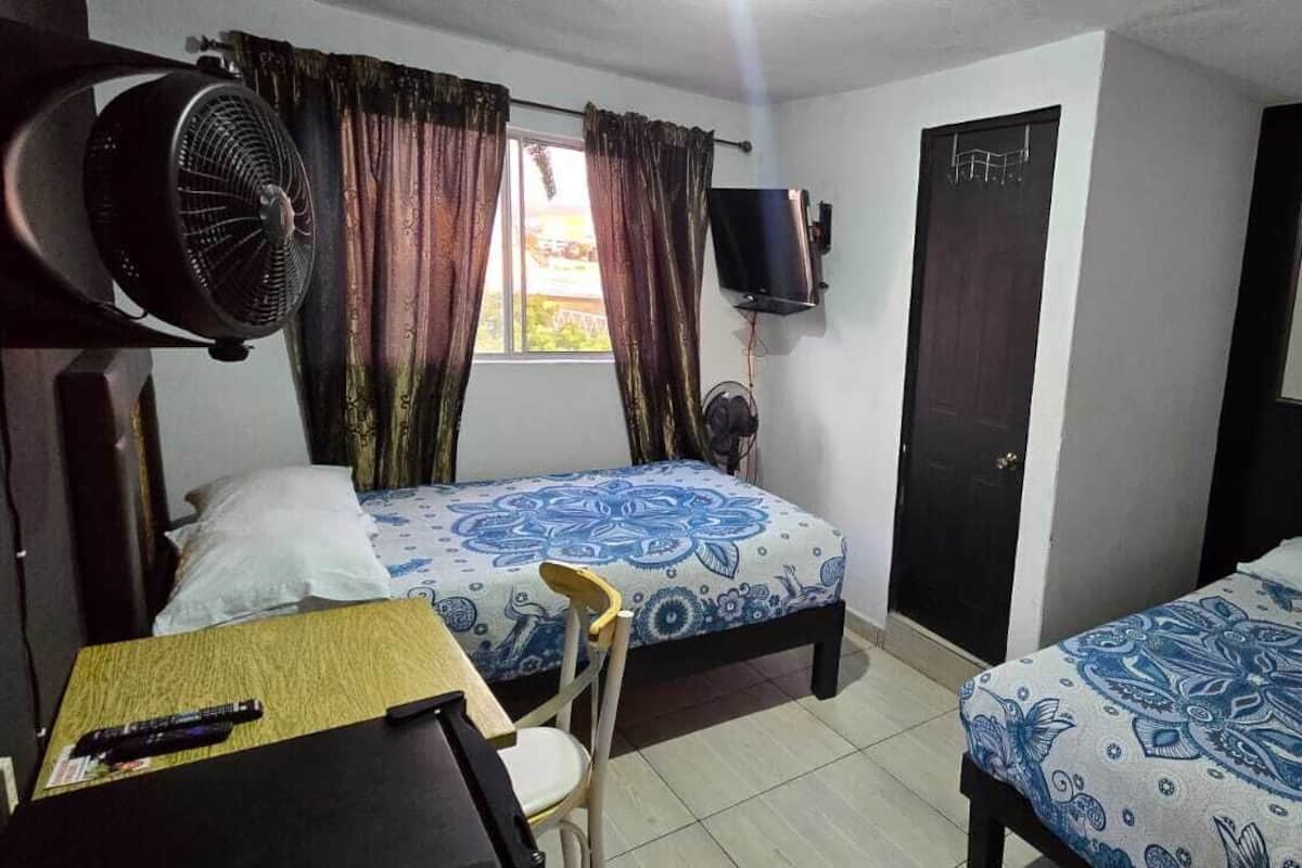 Quarto com duas camas, uma escrivaninha, uma cadeira, televisão e janela com cortinas.