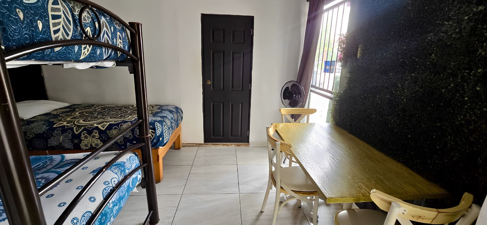 Quarto em dormitório com beliches, escrivaninha, cadeira, ventilador e porta.