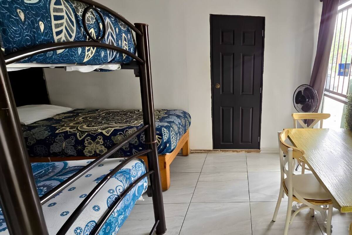 Quarto em dormitório com beliches, escrivaninha, cadeira, ventilador e porta.