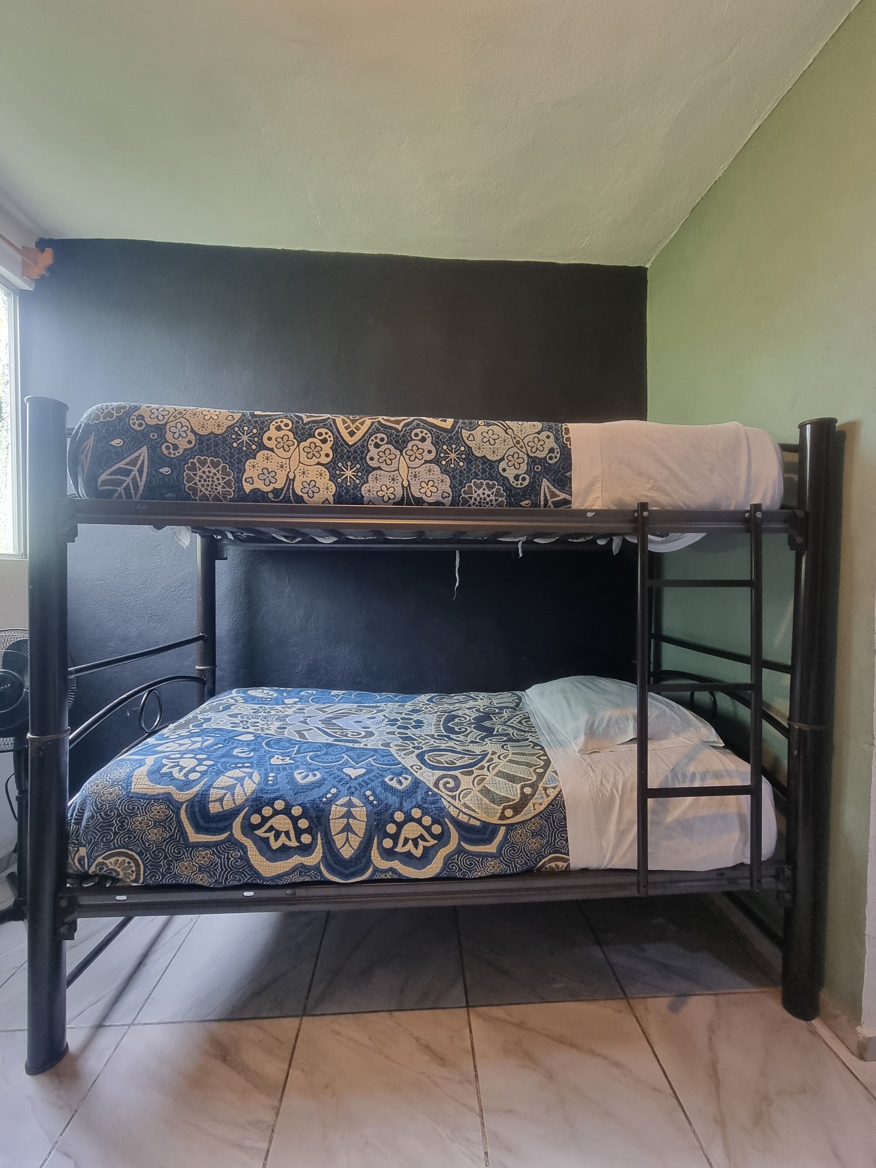 Uma cama beliche com roupa de cama estampada, um ventilador e uma janela num quarto com paredes verdes.