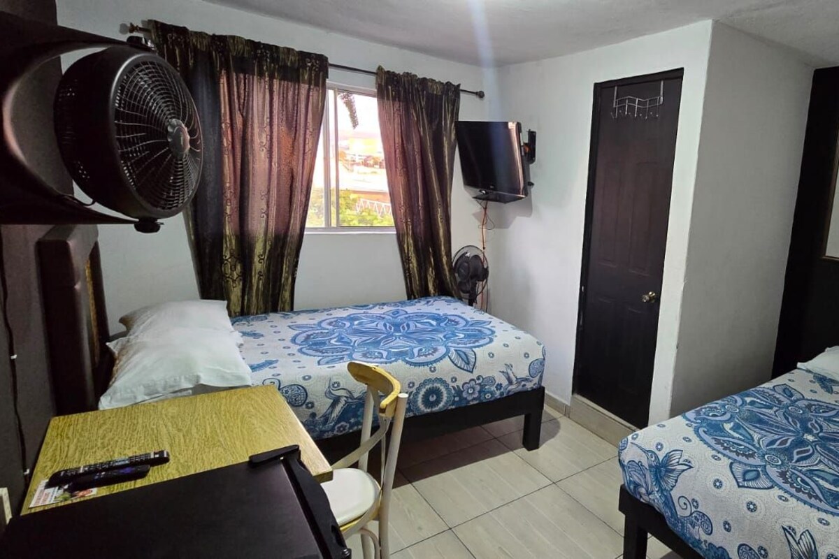 Quarto com duas camas, uma escrivaninha, uma cadeira, televisão e janela com cortinas.
