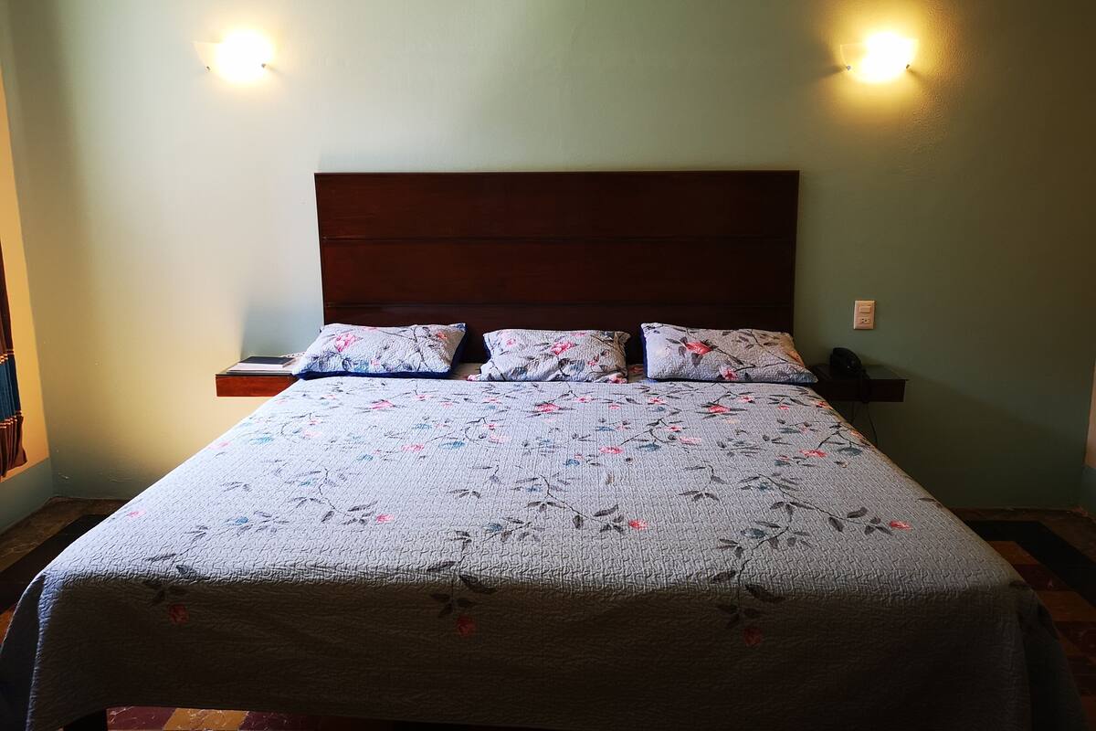 Cama com colcha florida, duas luminárias de apoio e mesa de cabeceira com telefone.