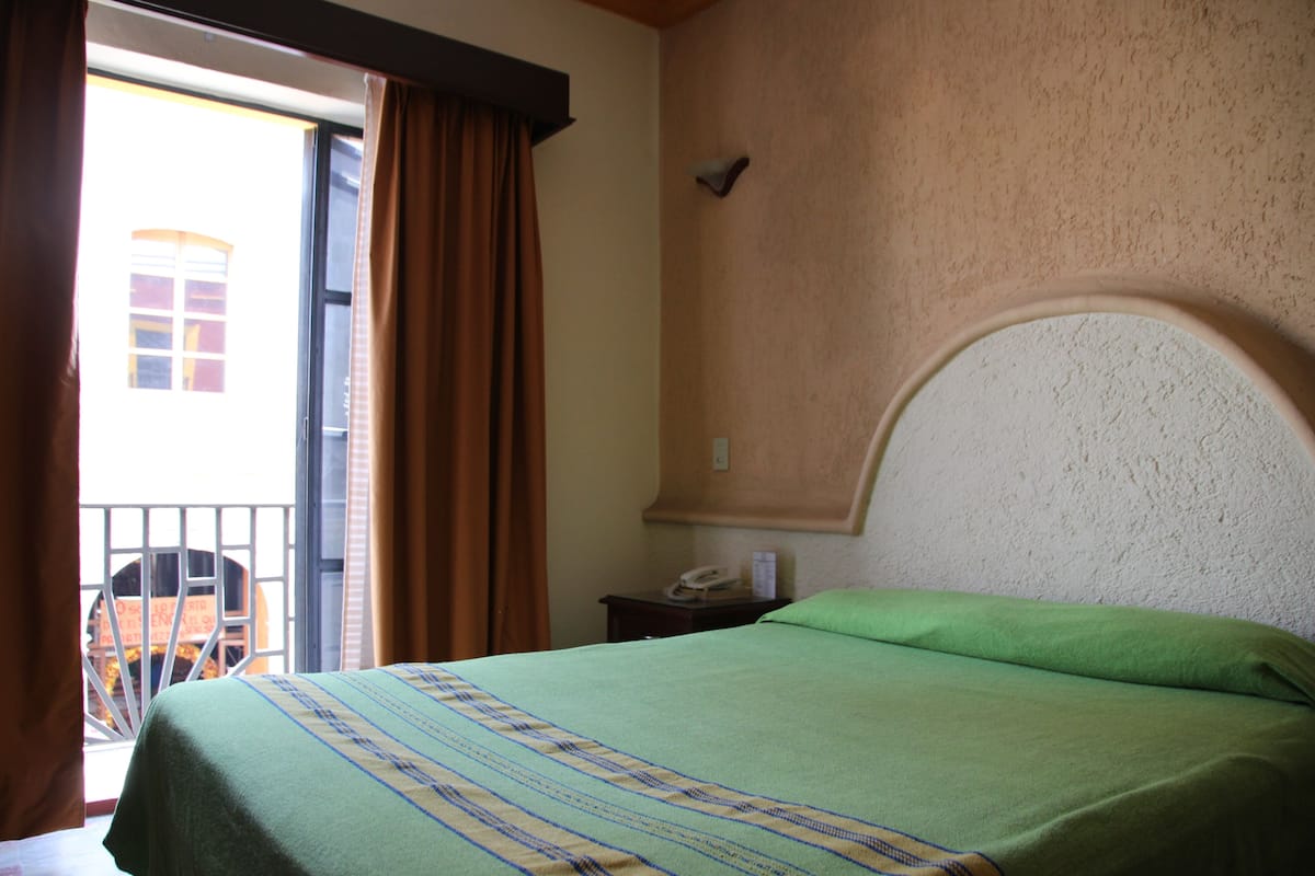 Quarto de hotel com uma cama, um telefone numa mesa de cabeceira, uma janela com cortinas e vista para o exterior.