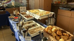 Uma mesa de buffet com diversos alimentos, utensílios, uma placa indicando a localização e um cesto de pão.