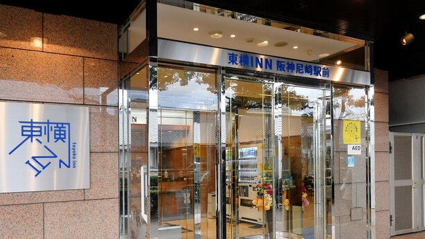 Entrada de hotel com porta de vidro, placa indicando "Toyoko Inn" e sinal de saída de emergência visível.