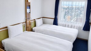 Quarto de hotel com duas camas, uma janela com cortinas, uma pequena prateleira com objetos e vista para a cidade.