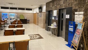 Um hall amplo com piso de azulejos, várias áreas de estar, máquinas de venda automática, uma pequena mesa com cartazes informativos e uma televisão na parede.
