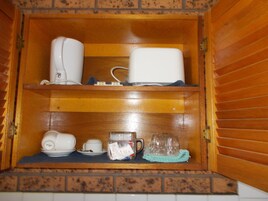 Um armário de madeira com duas prateleiras, contendo uma chaleira elétrica branca, uma cafeteira branca, um frasco de vidro com um sachê de chá, uma caneca de vidro e um pano verde dobrado.