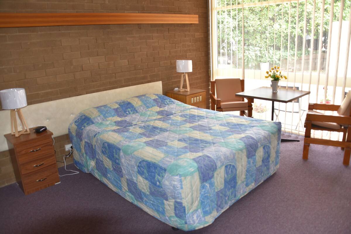Quarto com cama, escrivaninha, cadeira e janela com vista para o verde.