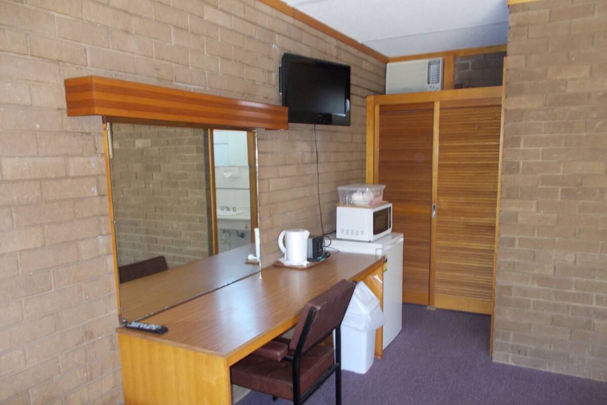 Quarto compacto com uma mesa de madeira, uma única cadeira marrom, micro-ondas e televisão fixada na parede.
