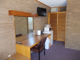 Quarto compacto com uma mesa de madeira, uma única cadeira marrom, micro-ondas e televisão fixada na parede.