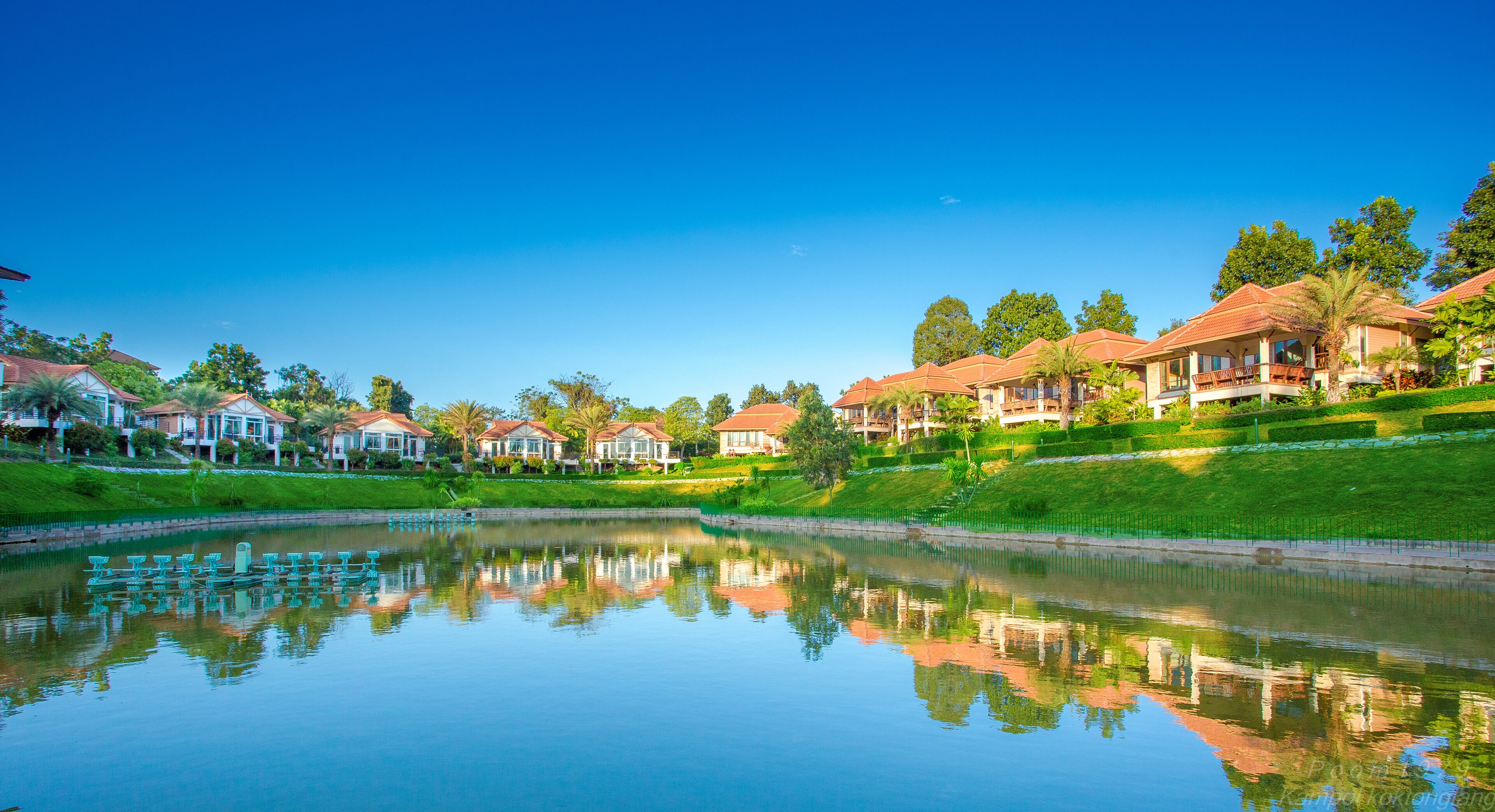 Vista serena de um lago, com várias casas, céu claro e águas calmas que refletem a paisagem.