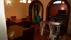 Uma área de refeições com mesas e cadeiras de madeira, uma pequena mesa branca com estrutura metálica e uma televisão ao fundo.