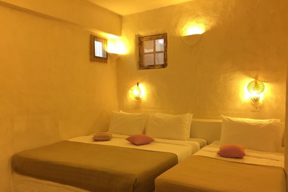 Quarto de hotel com duas camas, uma janela pequena e luminárias fixadas na parede.