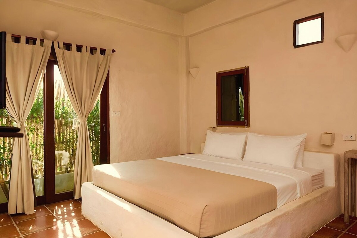 Quarto com cama, cortinas, uma pequena janela e vista para o verde lá fora.