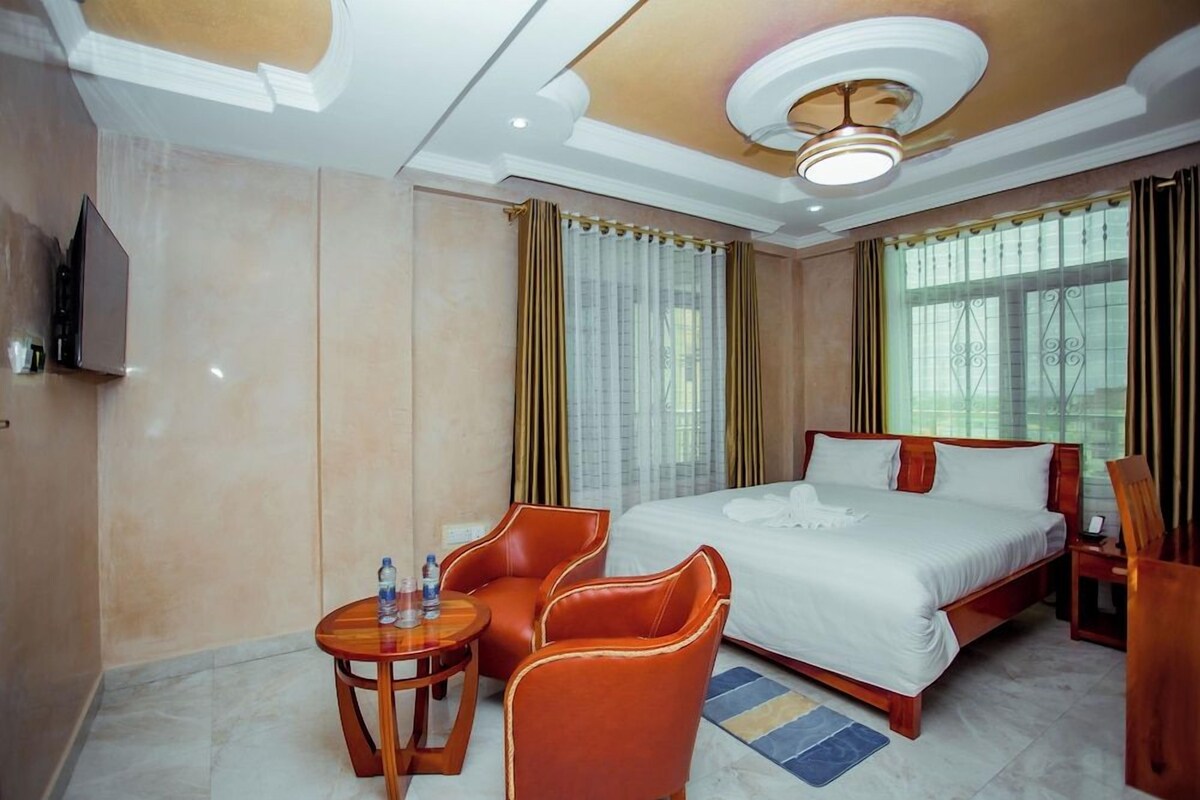Quarto de hotel com uma cama grande, duas poltronas laranja, uma mesinha com uma garrafa e uma televisão na parede.